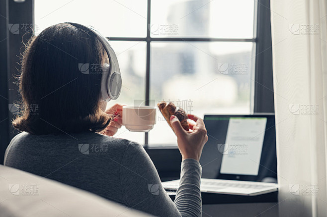 一个孤独的女人享受着一杯咖啡和羊角面包，戴着耳机听音乐，早上坐在窗边用笔记本电脑工作。