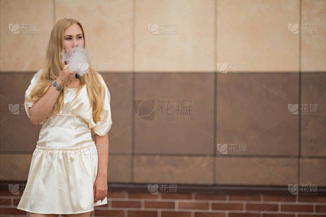 Vape 少年。一个年轻可爱的白色女孩穿着连衣裙是 vaping 一根电子香烟对面的墙上的街道上的夏