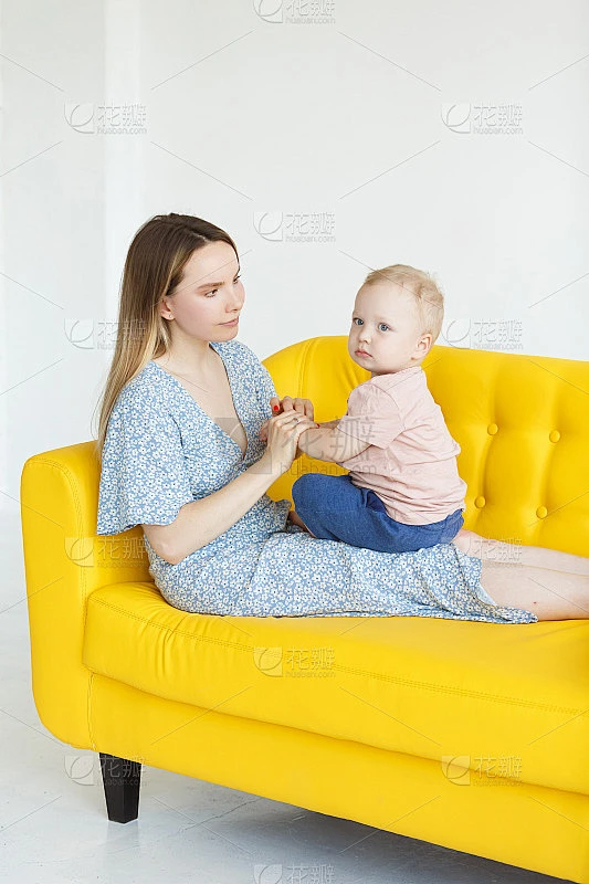 快乐的年轻妈妈和她可爱的小儿子一起玩在明亮的黄色沙发在白色的室内，微笑的妈妈和孩子手牵手，一起玩，垂素材 花瓣网eow 5461