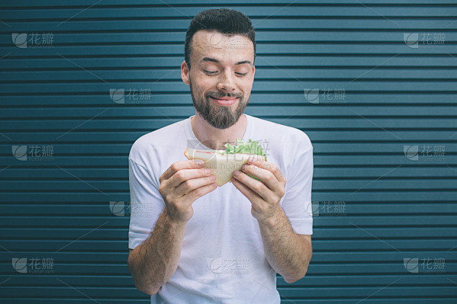 快乐的家伙手里拿着三明治，看着它。他是面带微笑。孤立在条纹和蓝色背景。