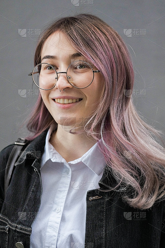 一个穿着牛仔夹克，戴着眼镜的年轻女人。潮人紫色的头，在灰色的墙壁背景上戴着圆眼镜。